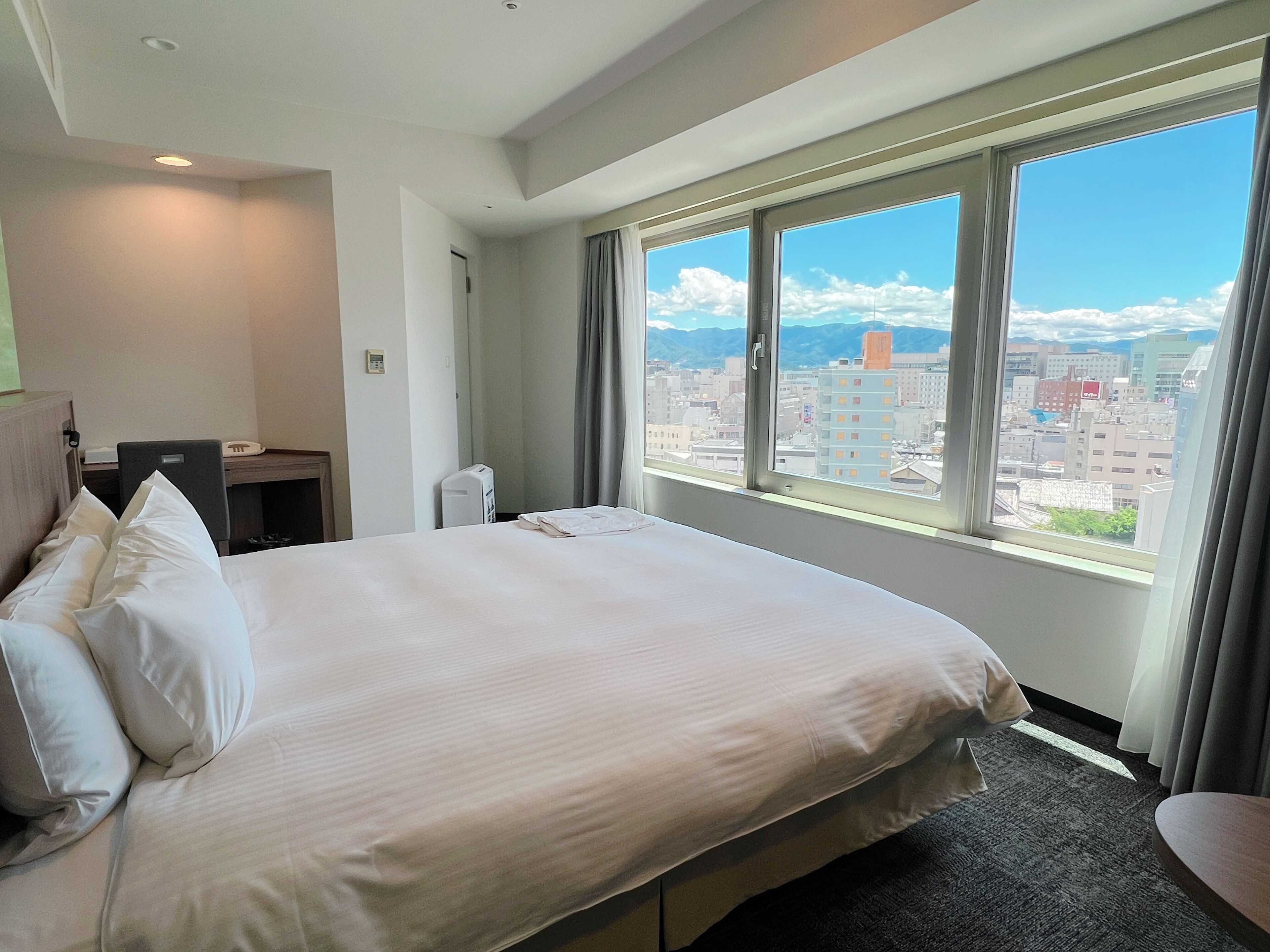 Quarto de hotel com uma cama grande, uma mesa de trabalho, uma cadeira e vista para a cidade através de amplas janelas.