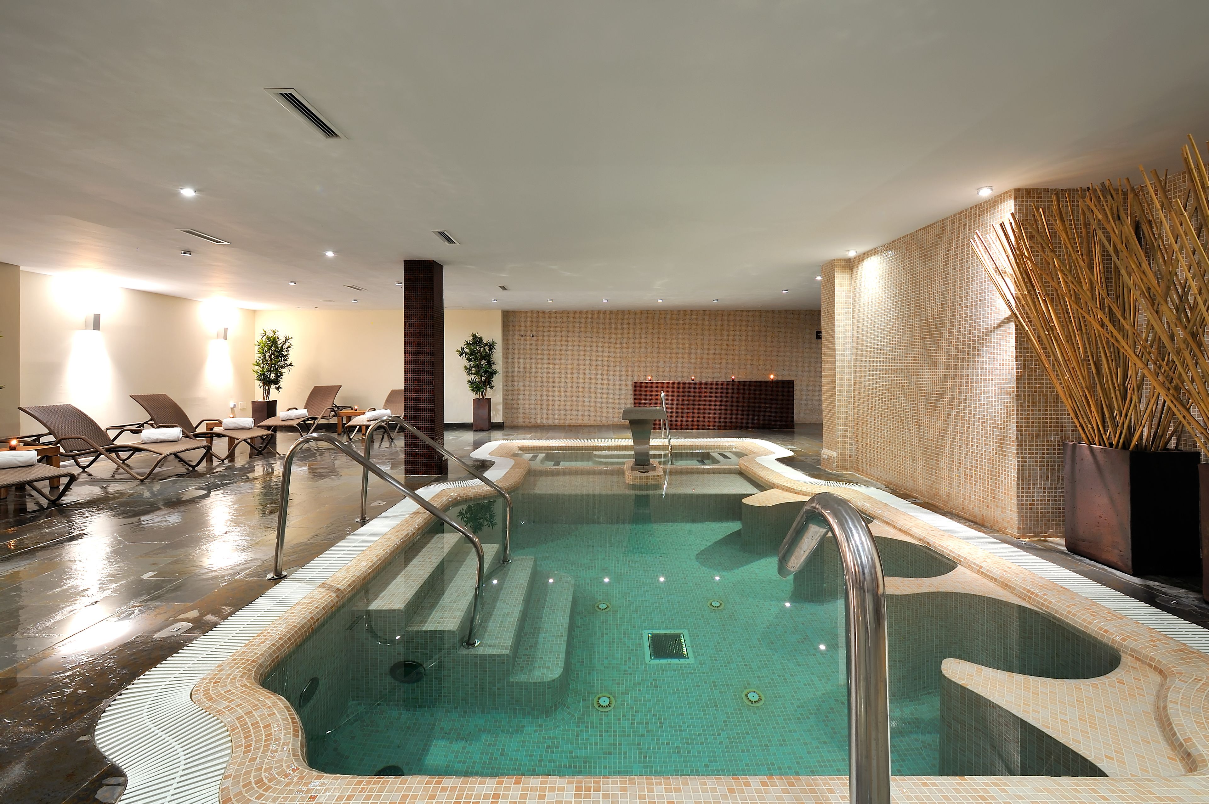Uma grande piscina interior com borda revestida em azulejo, uma área de recepção com balcão e cadeiras, e uma parede decorada com bambu.