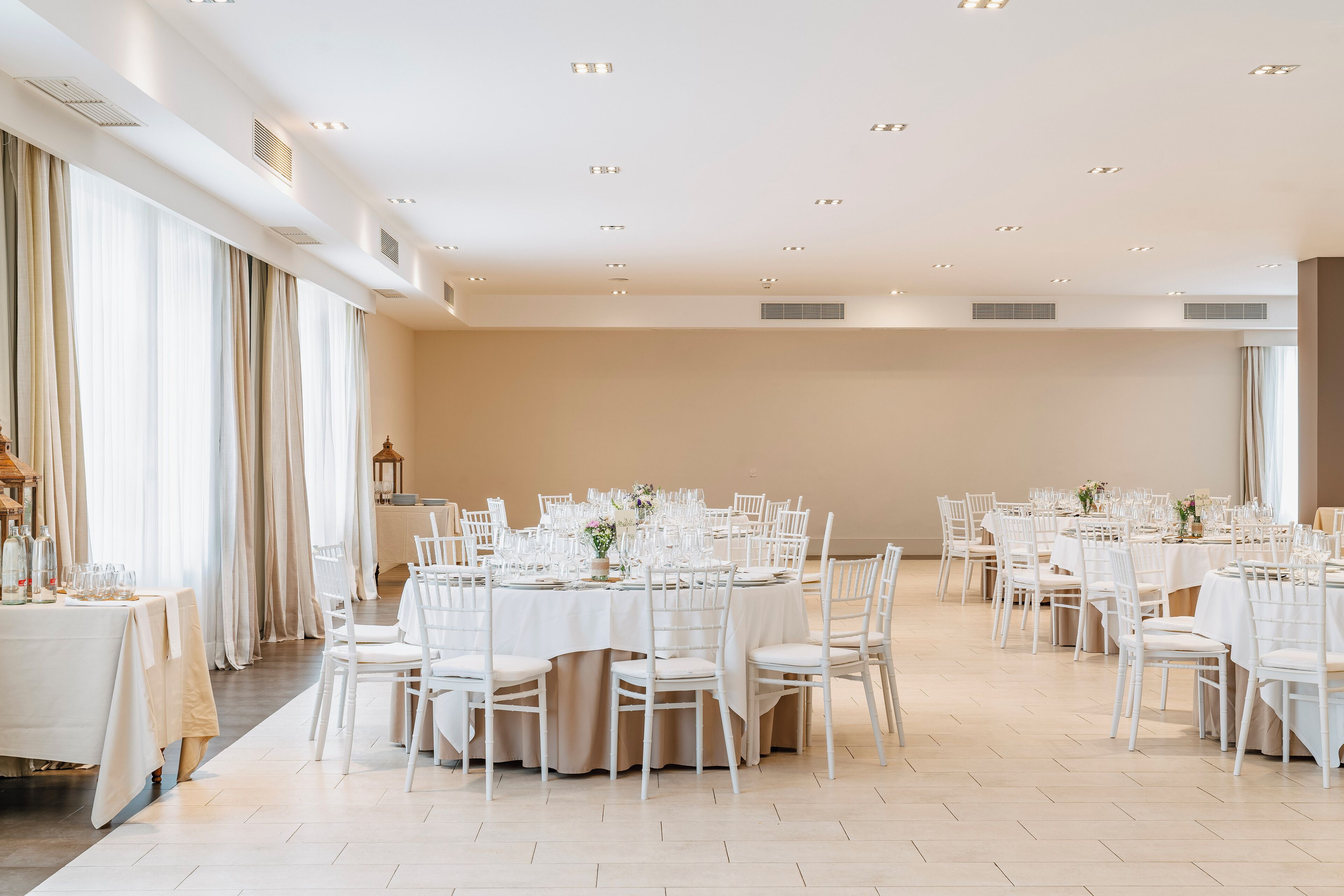 Uma sala de banquetes ampla, com várias mesas redondas, cada uma com toalhas brancas e cadeiras, e uma paleta de cores neutras.
