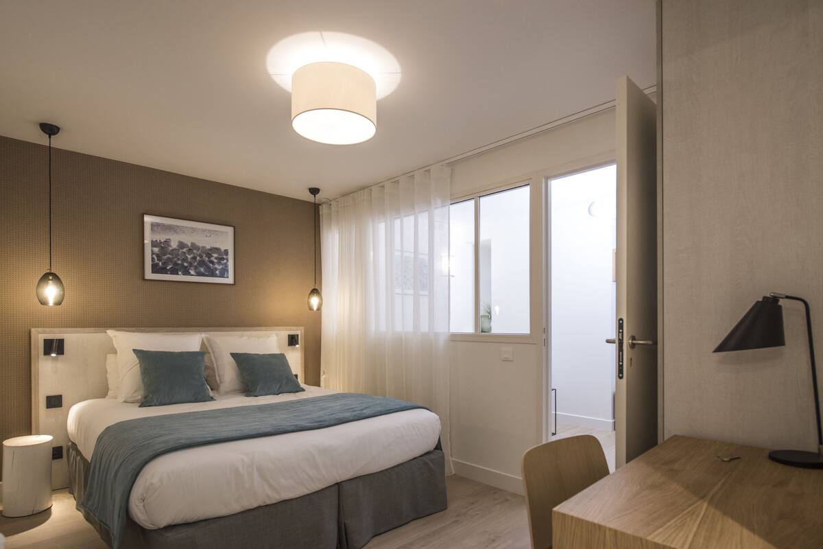 Quarto de hotel moderno com uma cama grande, luminárias laterais, uma escrivaninha e vista da cidade através de janelas amplas.