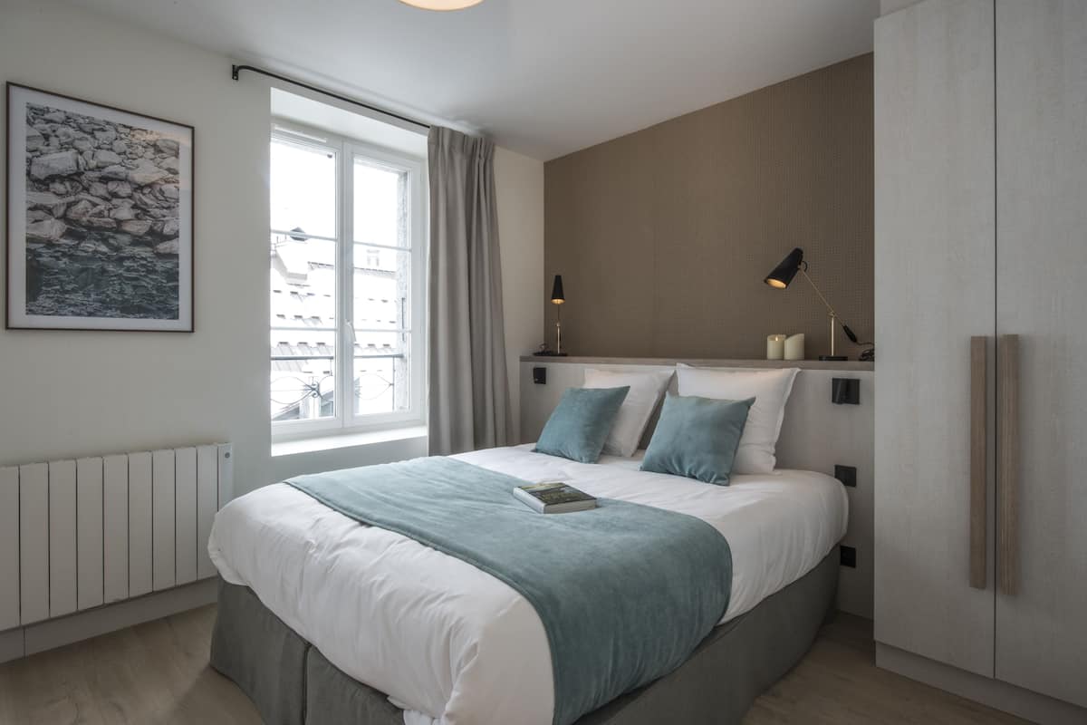 Quarto moderno com uma cama grande, janela com cortinas, radiador e um quadro na parede.