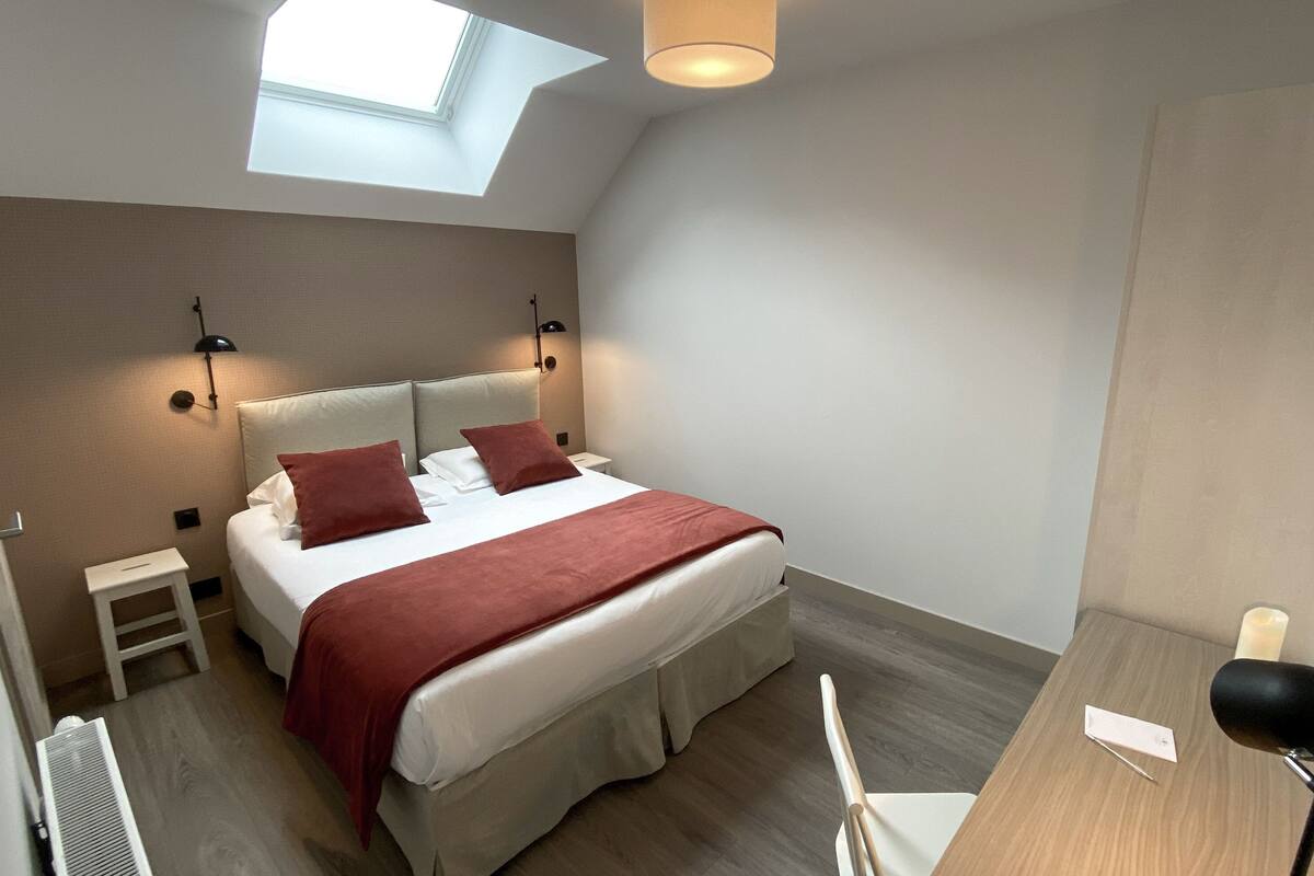 Um quarto moderno com claraboia, uma cama com manta vermelha, mesas de cabeceira e uma escrivaninha com cadeira.