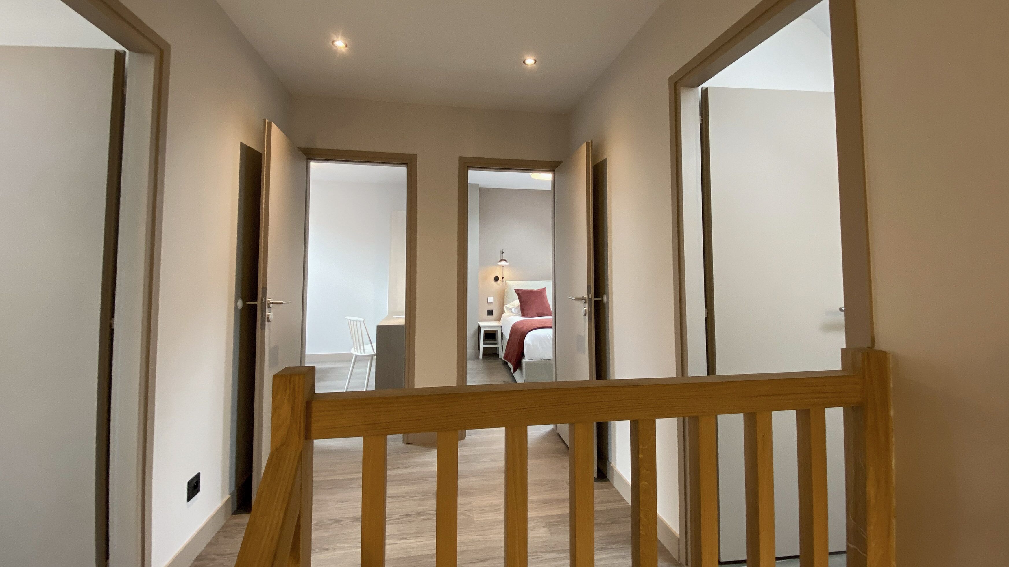 Um corredor com piso e corrimão de madeira. Há portas que dão para um quarto com cama, mesa de cabeceira e uma cadeira.