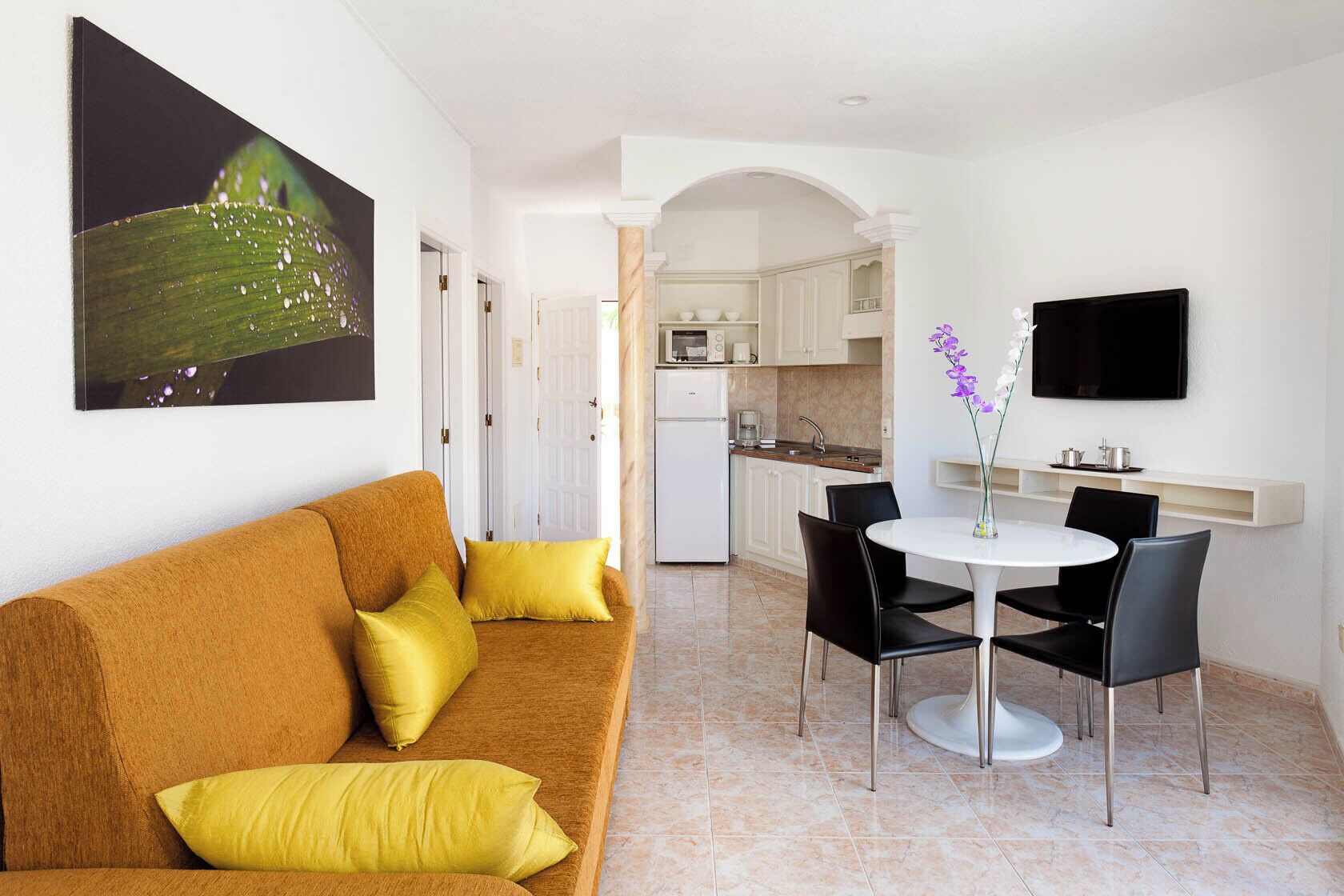 Uma sala de estar moderna com um sofá marrom e almofadas amarelas, uma área de jantar com uma mesa branca e cadeiras pretas, uma cozinha com armários brancos e uma TV de tela plana fixada na parede.