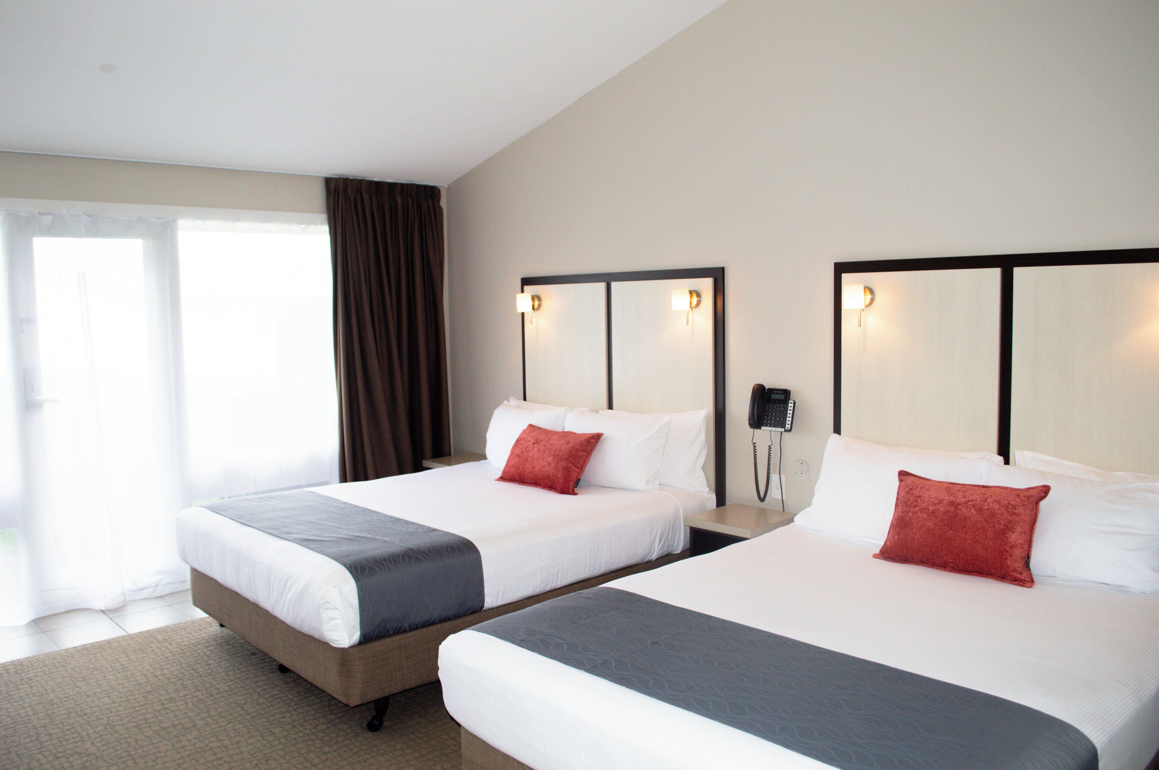 Quarto de hotel com duas camas, uma janela grande com cortinas e um telefone na parede.