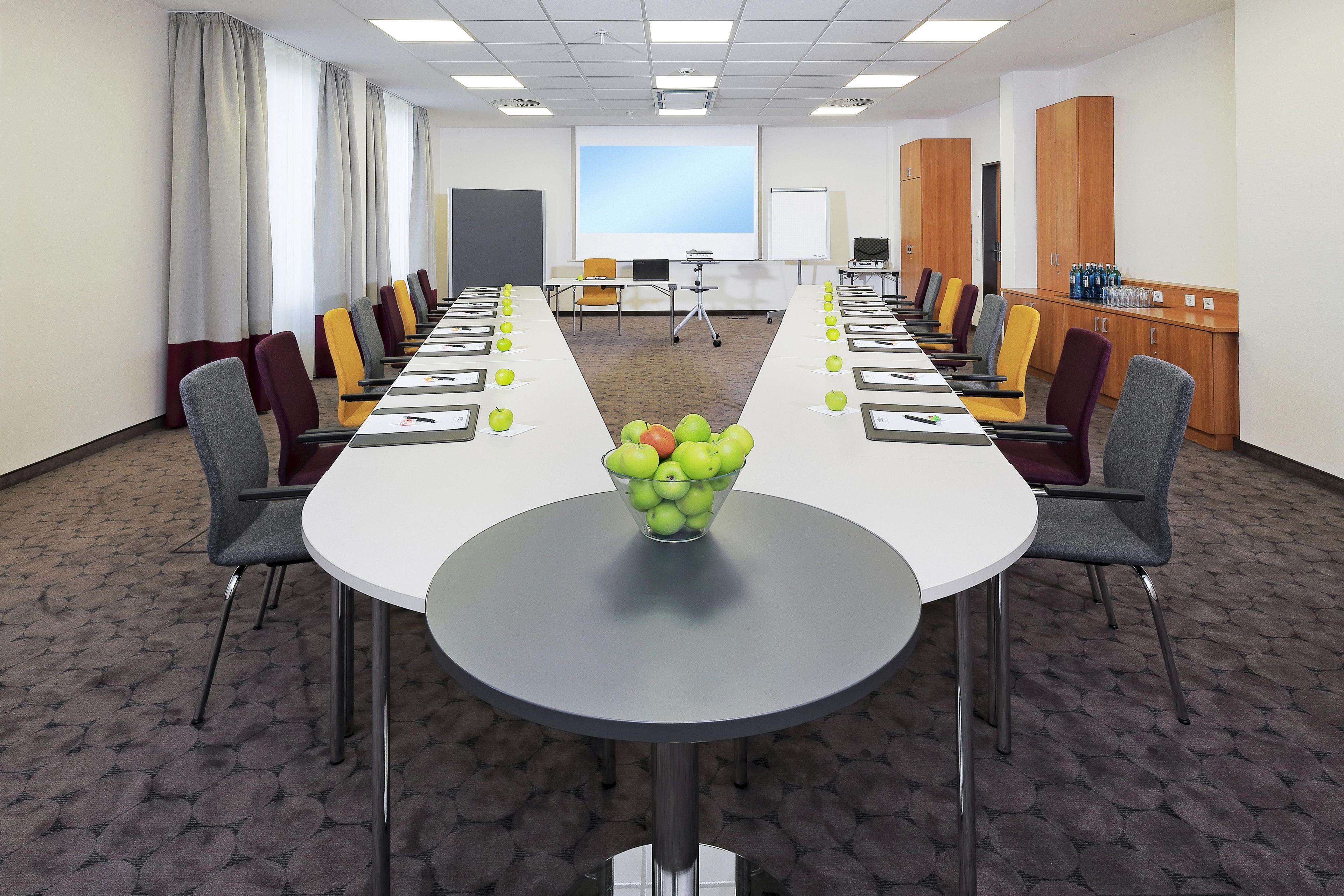 Sala de conferência com uma mesa grande, cadeiras, tela de projeção e bebedouro.