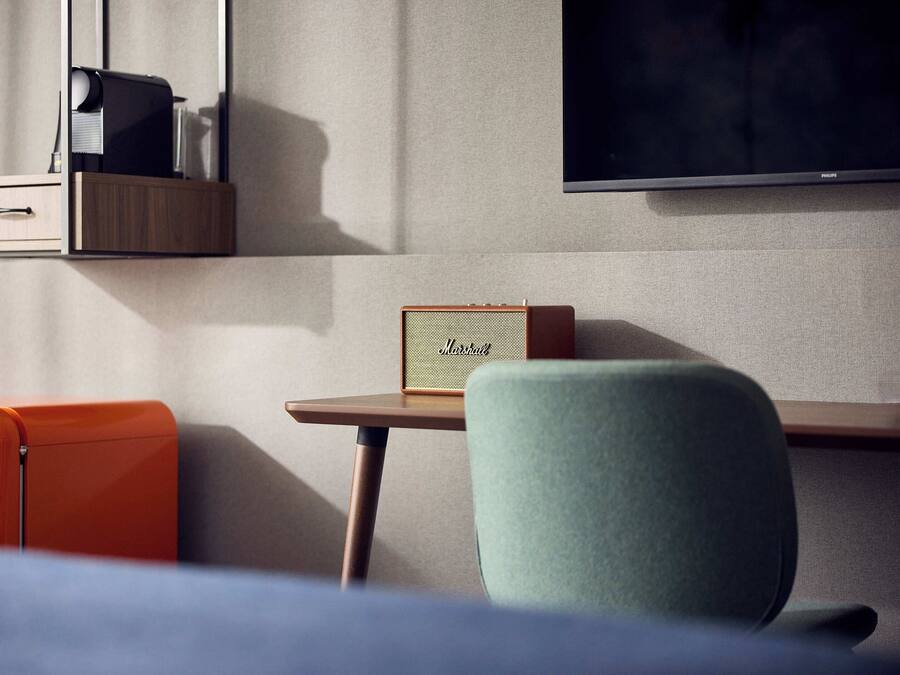 Quarto de hotel moderno com televisão de tela plana na parede, pequena mesa de madeira, poltrona verde e unidade de armazenamento para malas laranja.