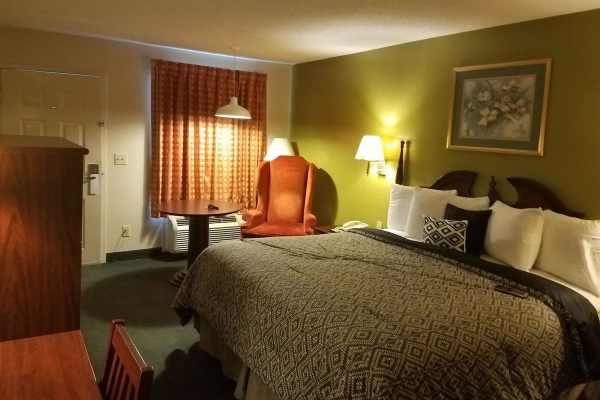 Quarto de hotel com cama, poltrona laranja, mesa, luminária e um quadro na parede.