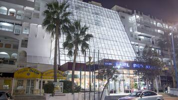 Um hotel com fachada de vidro moderna, palmeiras e um carro estacionado na frente.