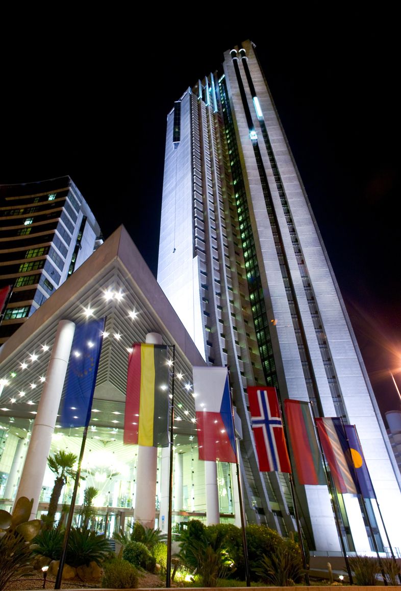 Um edifício moderno com várias bandeiras, incluindo a bandeira da União Europeia, e uma área de entrada bem iluminada com plantas.