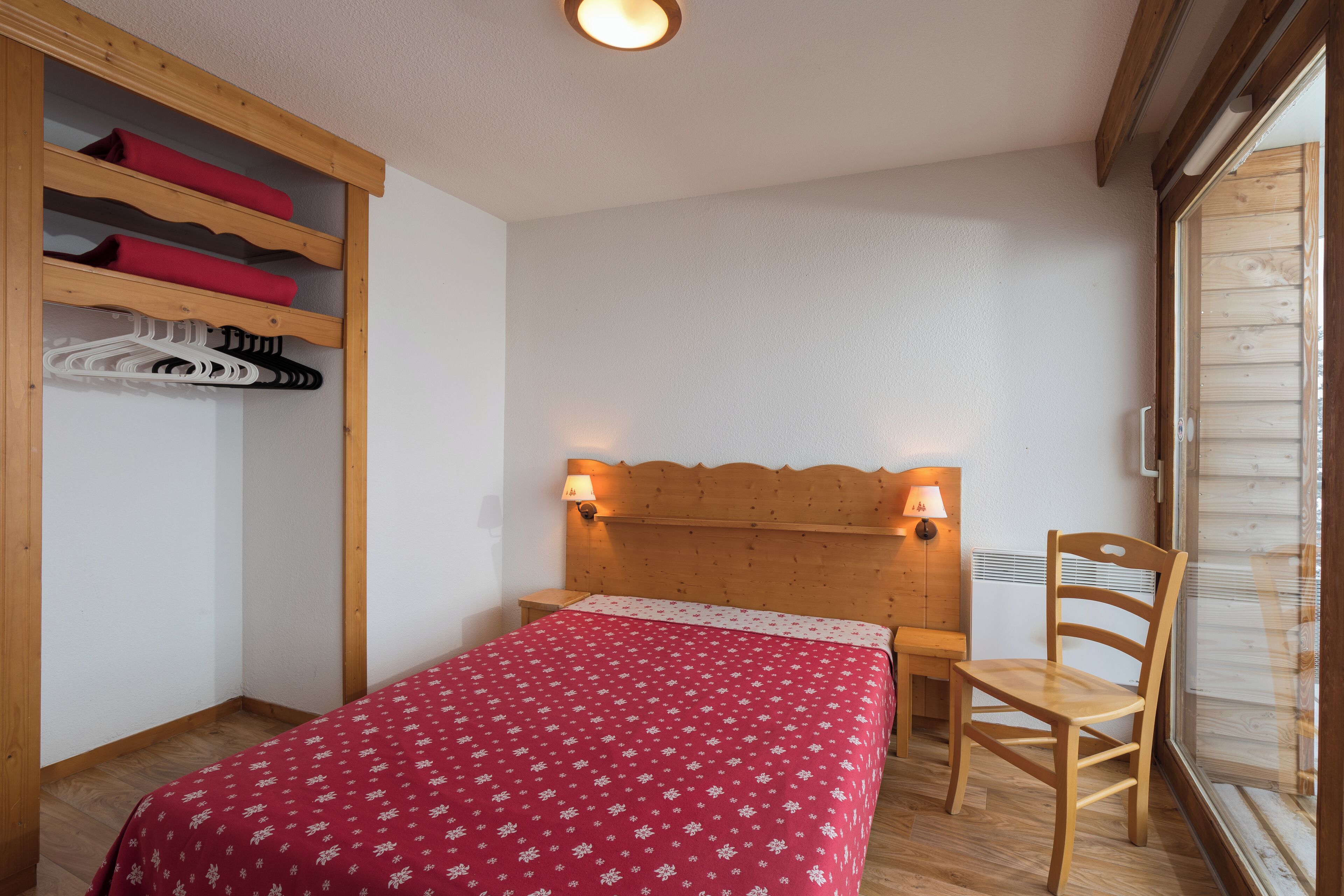 Quarto com cama de madeira, uma cadeira, mesa de cabeceira, guarda-roupa com prateleiras e uma janela.