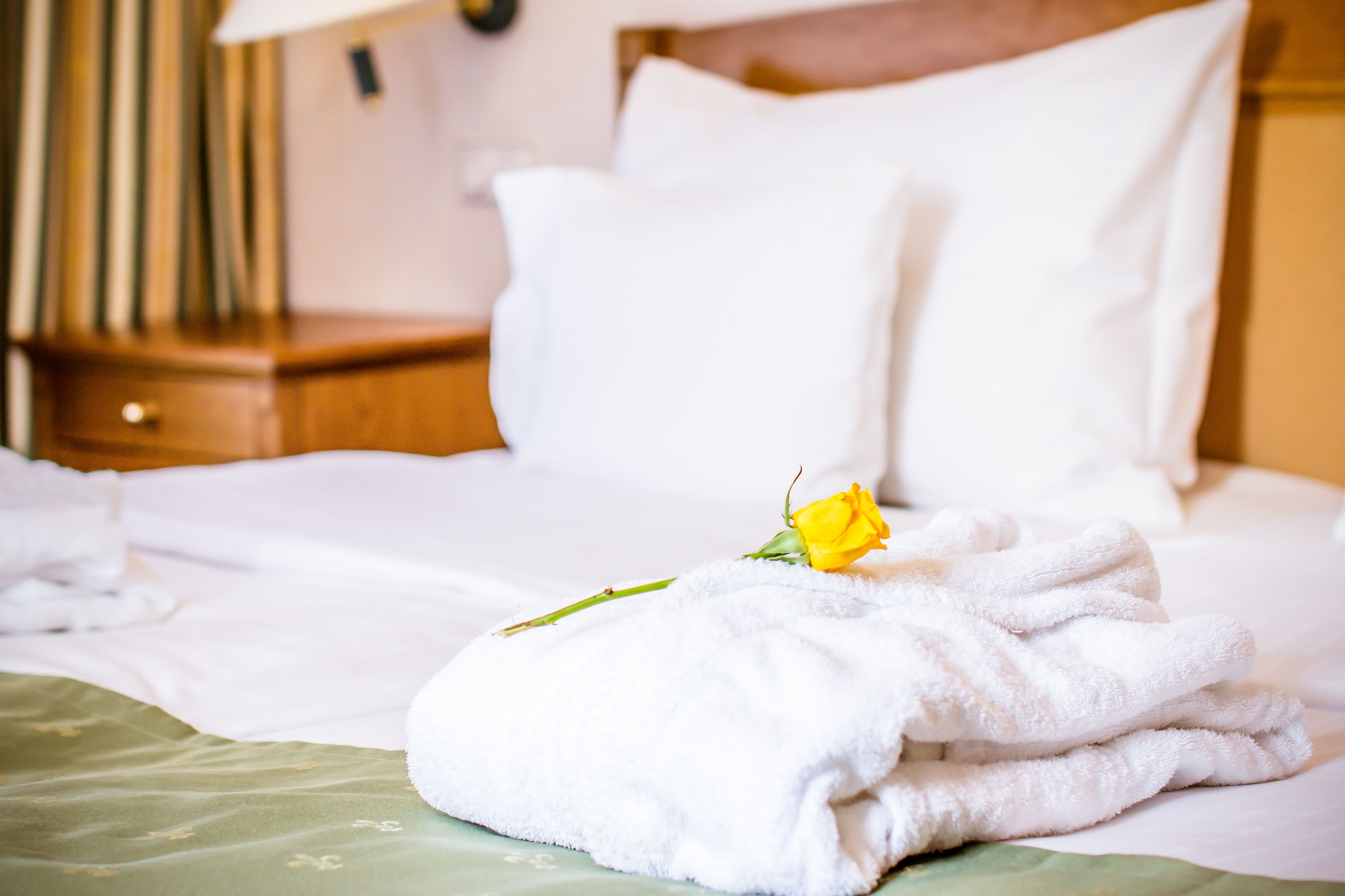 Uma cama bem arrumada com lençóis brancos, uma única rosa amarela e uma toalha branca dobrada.