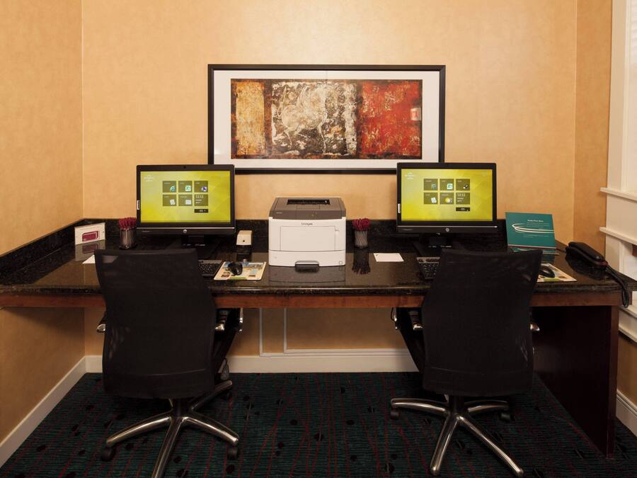Quarto de hotel com escrivaninha, duas cadeiras, dois laptops, uma impressora e uma obra de arte emoldurada na parede.