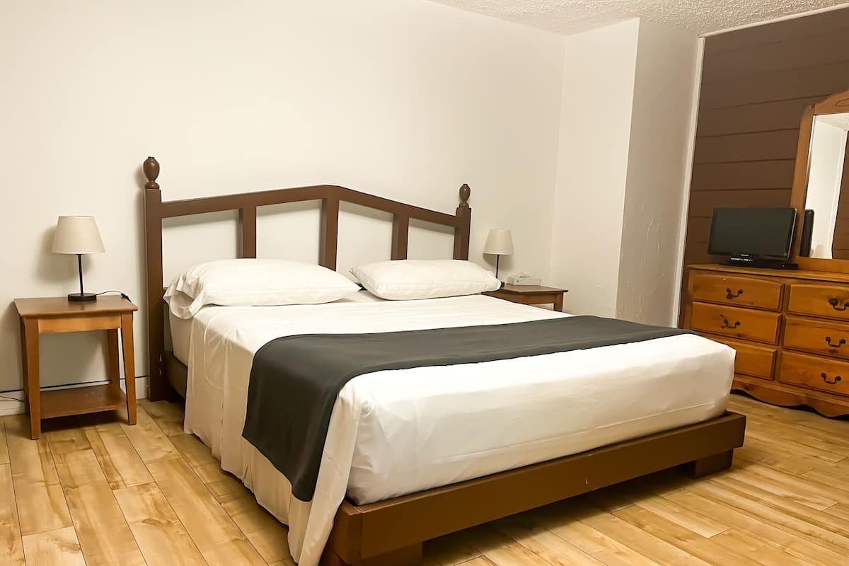 Cama bem arrumada com cabeceira de madeira, duas mesas de cabeceira com abajures e cômoda de madeira com televisão.