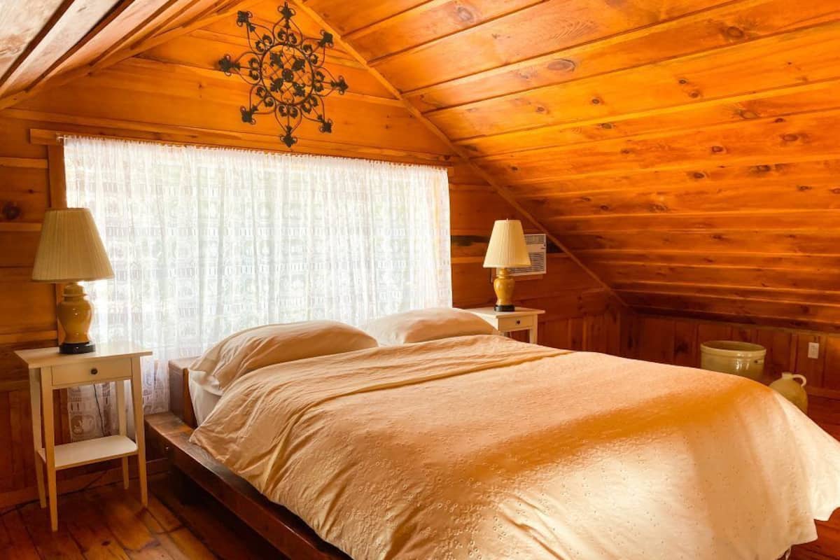 Quarto de sótão aconchegante com teto de madeira, uma cama grande com edredão branco, duas luminárias de cabeceira e um objeto decorativo na parede.
