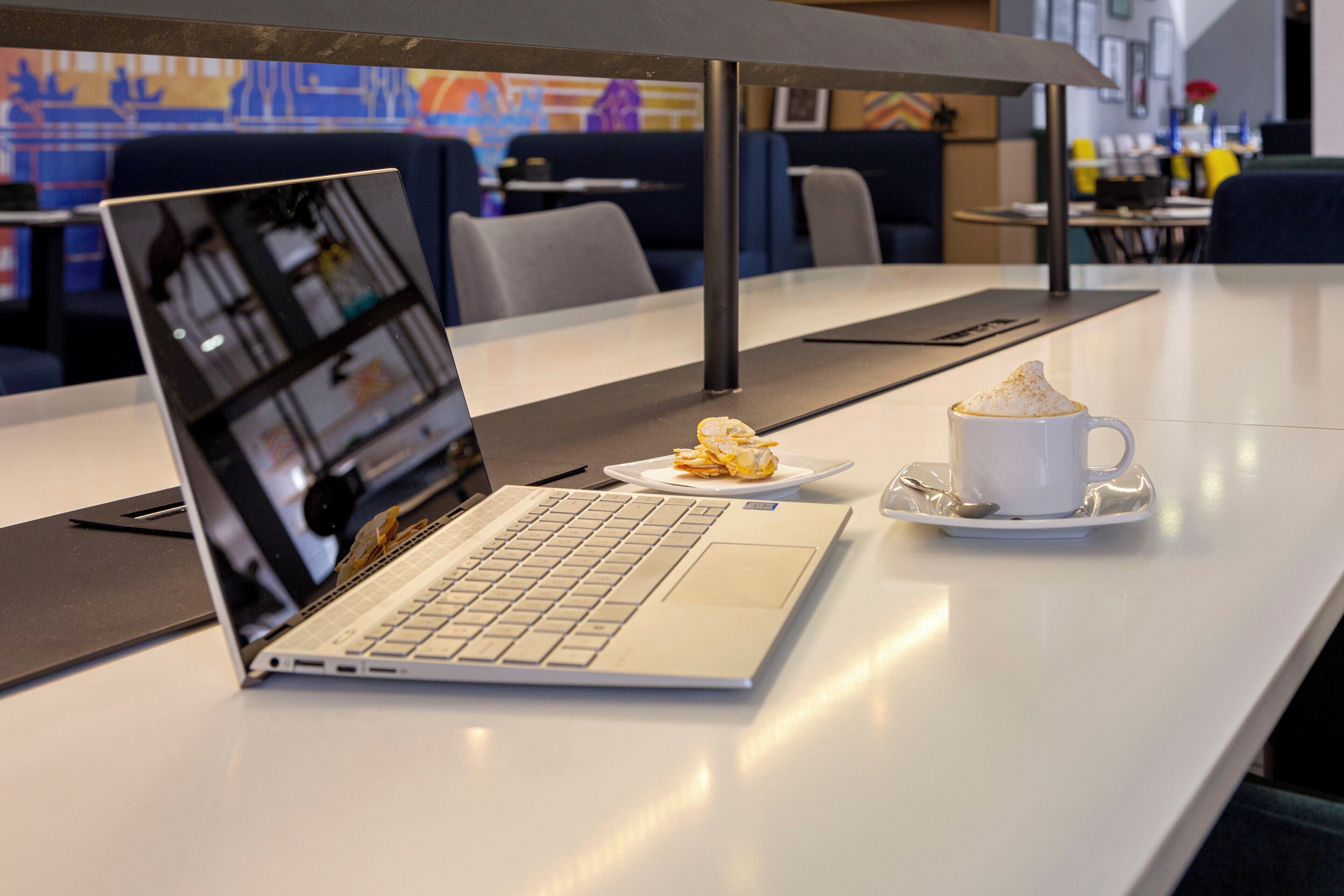 Um laptop sobre uma mesa, com uma xícara de café e um prato de doces.