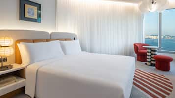 Quarto de hotel com uma cama grande, uma mesa de cabeceira com abajur, uma pequena mesa com cadeiras e vista para a cidade pela janela.