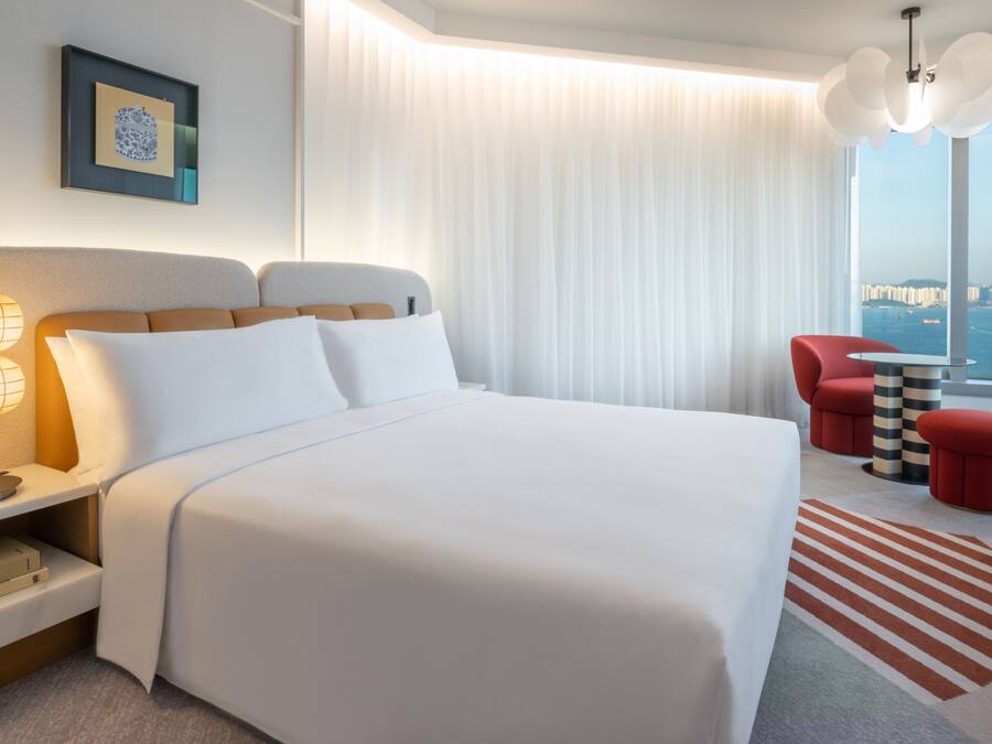Quarto de hotel com uma cama grande, uma mesa de cabeceira com abajur, uma pequena mesa com cadeiras e vista para a cidade pela janela.