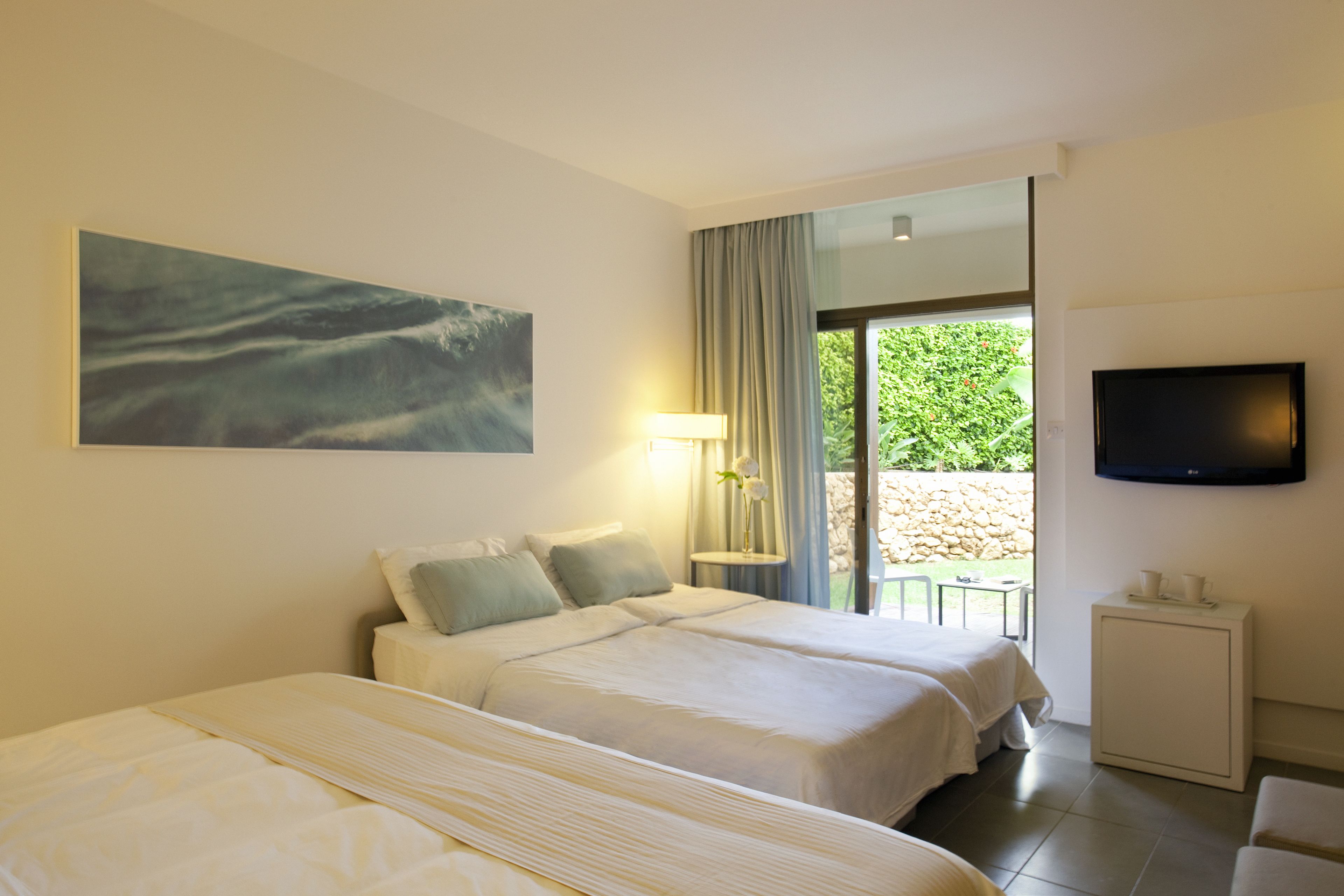 Quarto de hotel com duas camas, televisão, um quadro e varanda com vista para o verde.