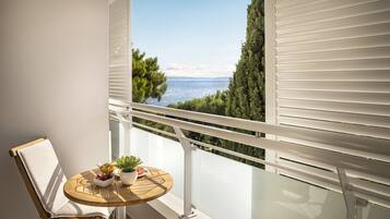 Uma varanda com vista para o mar, uma mesa de madeira redonda com uma cesta de frutas e um vaso de planta, e uma cadeira com almofada branca.