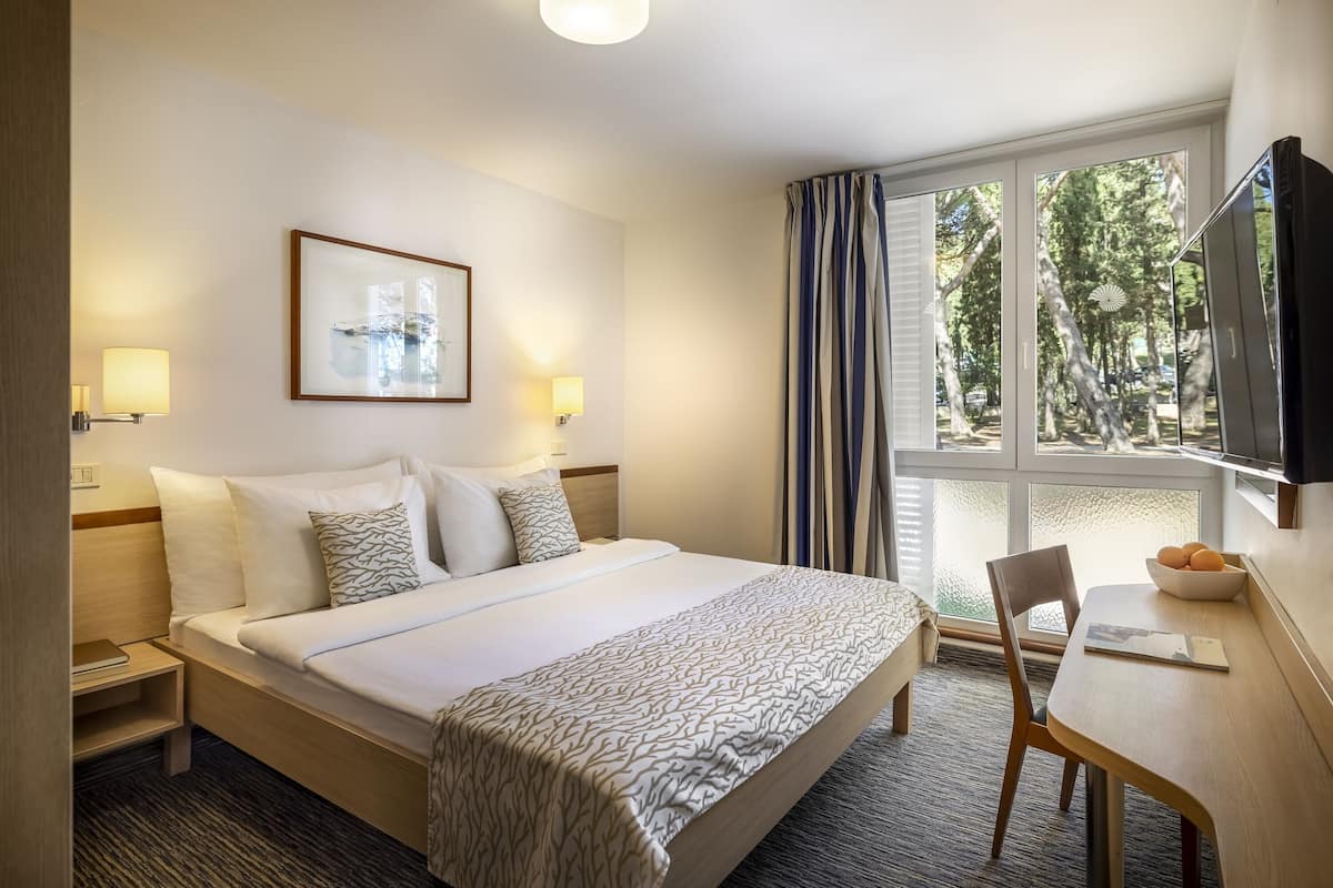 Quarto de hotel com uma cama grande, abajures, um quadro emoldurado, uma escrivaninha com uma tigela de laranjas e uma janela com cortinas.