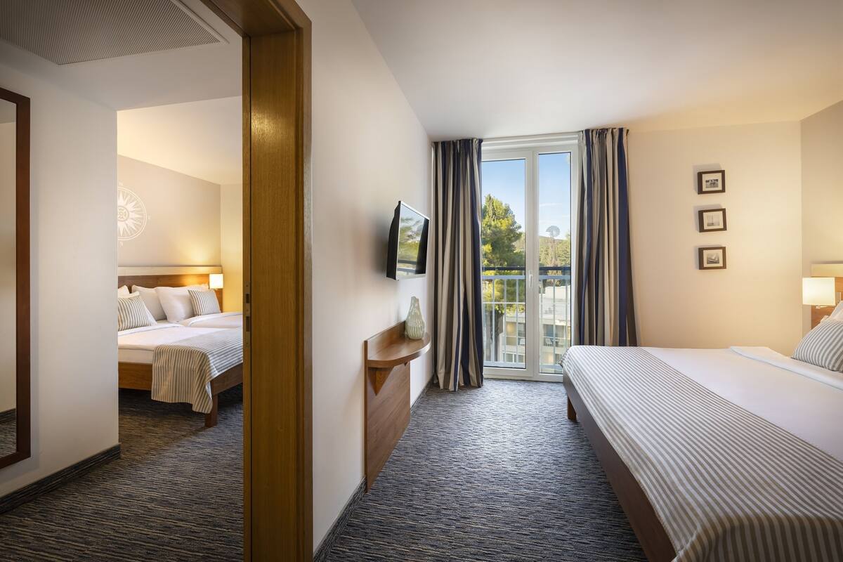 Quarto de hotel com cama, televisão, uma mesinha e vista para o exterior através de uma porta de vidro.