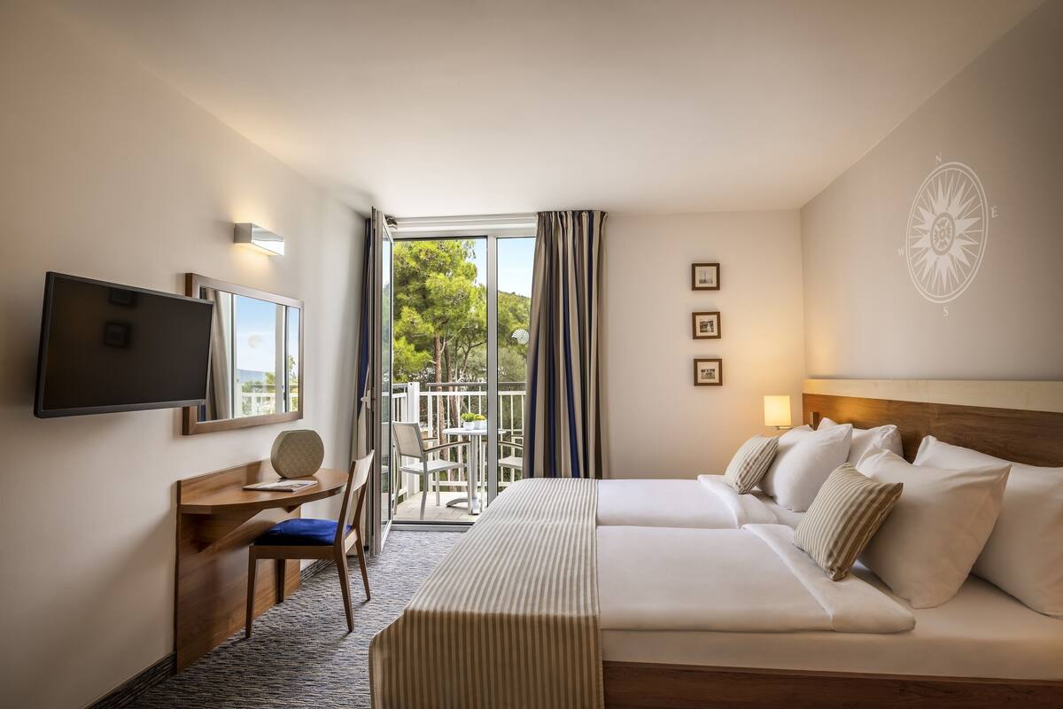 Quarto de hotel com cama, escrivaninha, cadeira, televisão e varanda com cortinas.
