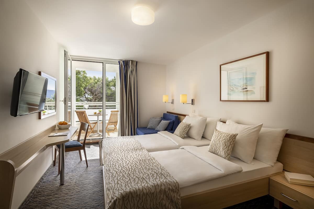 Quarto de hotel com duas camas, uma escrivaninha com televisão, zona de refeições com mesa e cadeiras e varanda com vista.