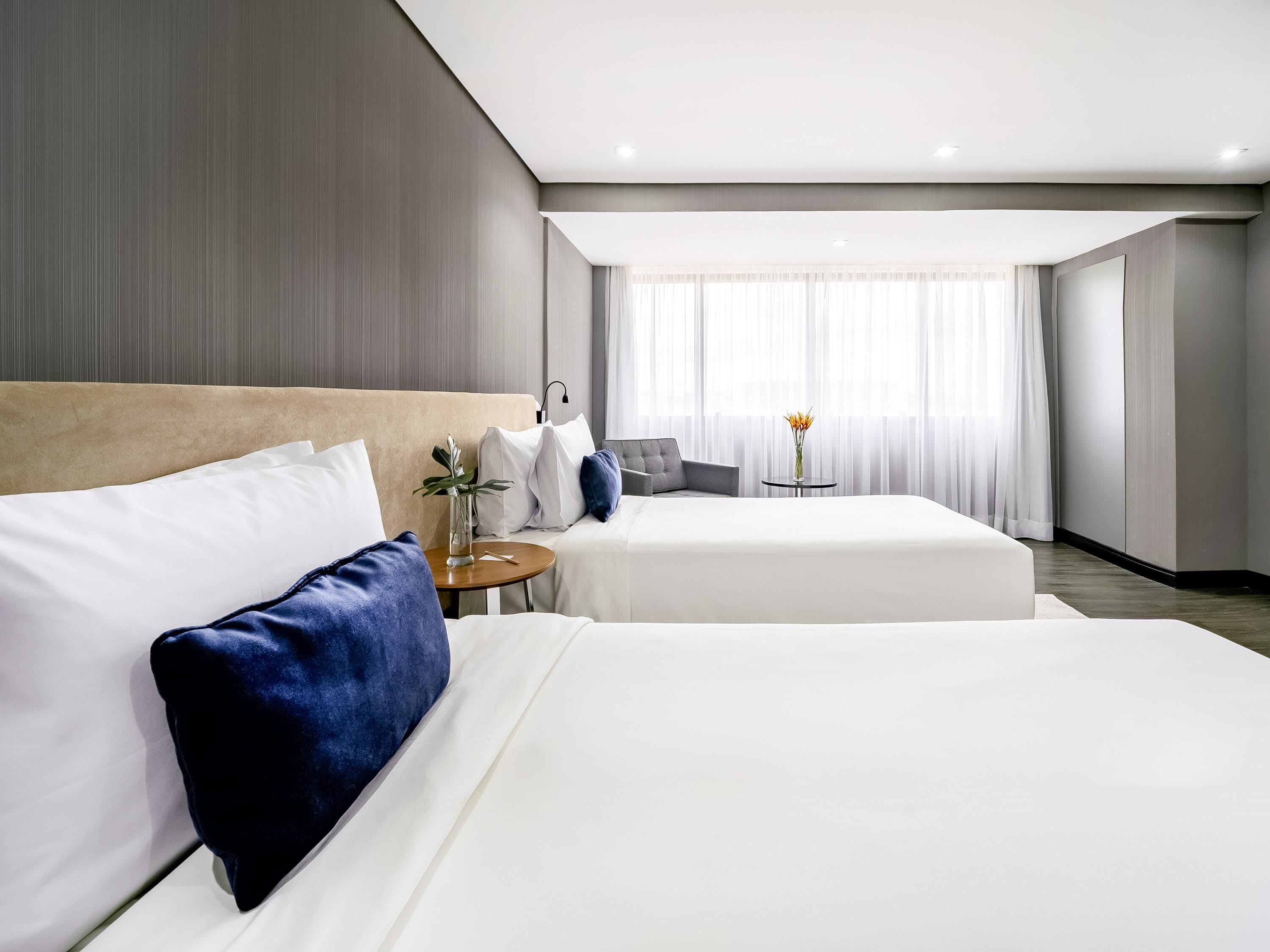 Quarto de hotel com duas camas, uma mesinha com um vaso e uma cadeira.