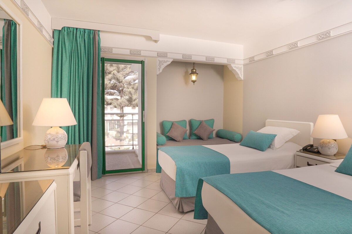 Quarto de hotel com duas camas, uma escrivaninha, televisão e varanda com vista.