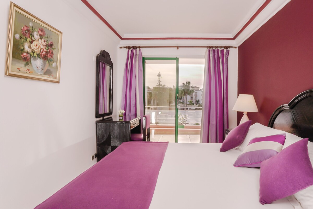 Quarto com cama, escrivaninha, cadeira, espelho, janela com cortinas e um quadro na parede.