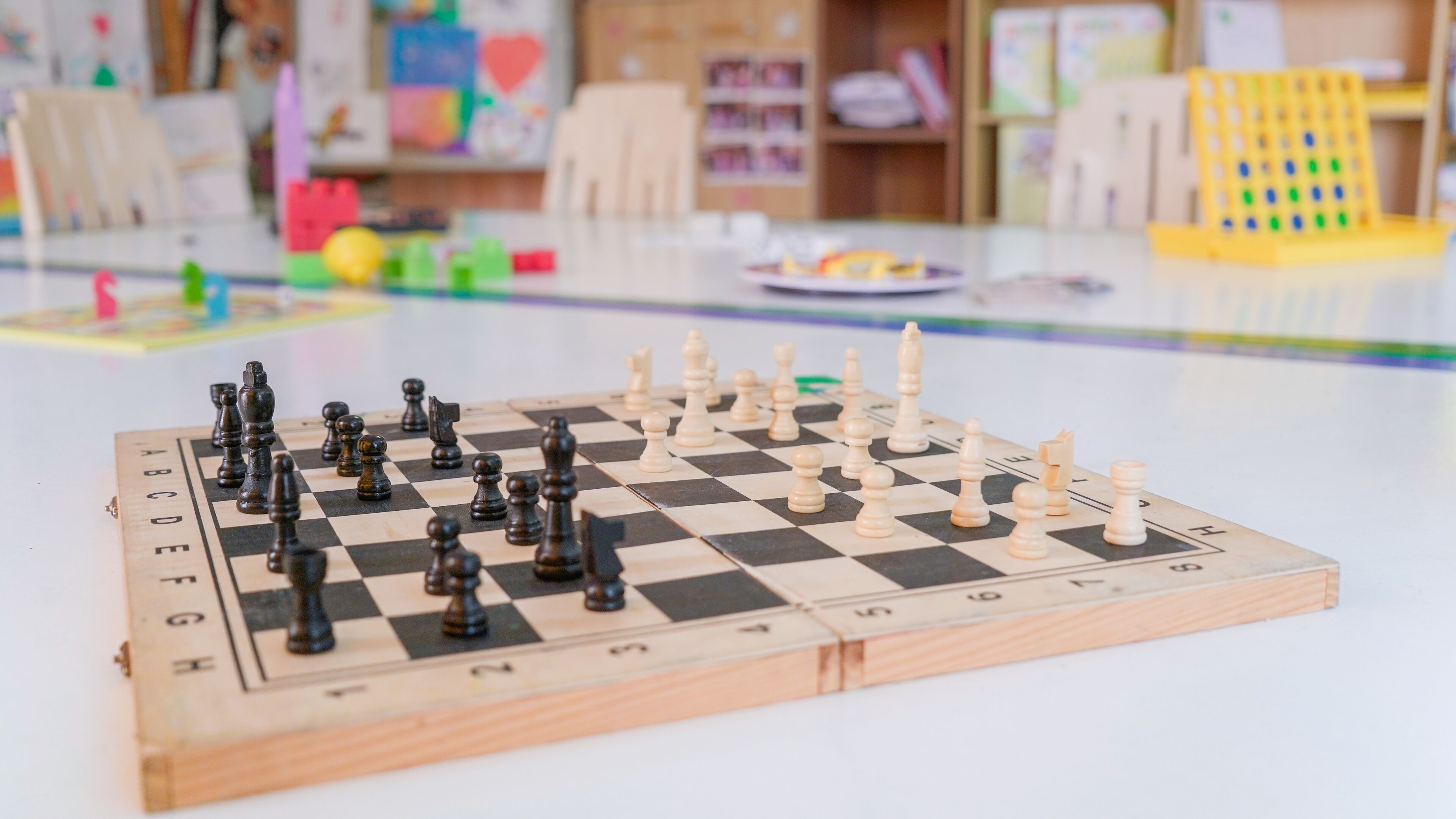 Um tabuleiro de xadrez com peças sobre uma mesa em um quarto com estantes e brinquedos ao fundo.