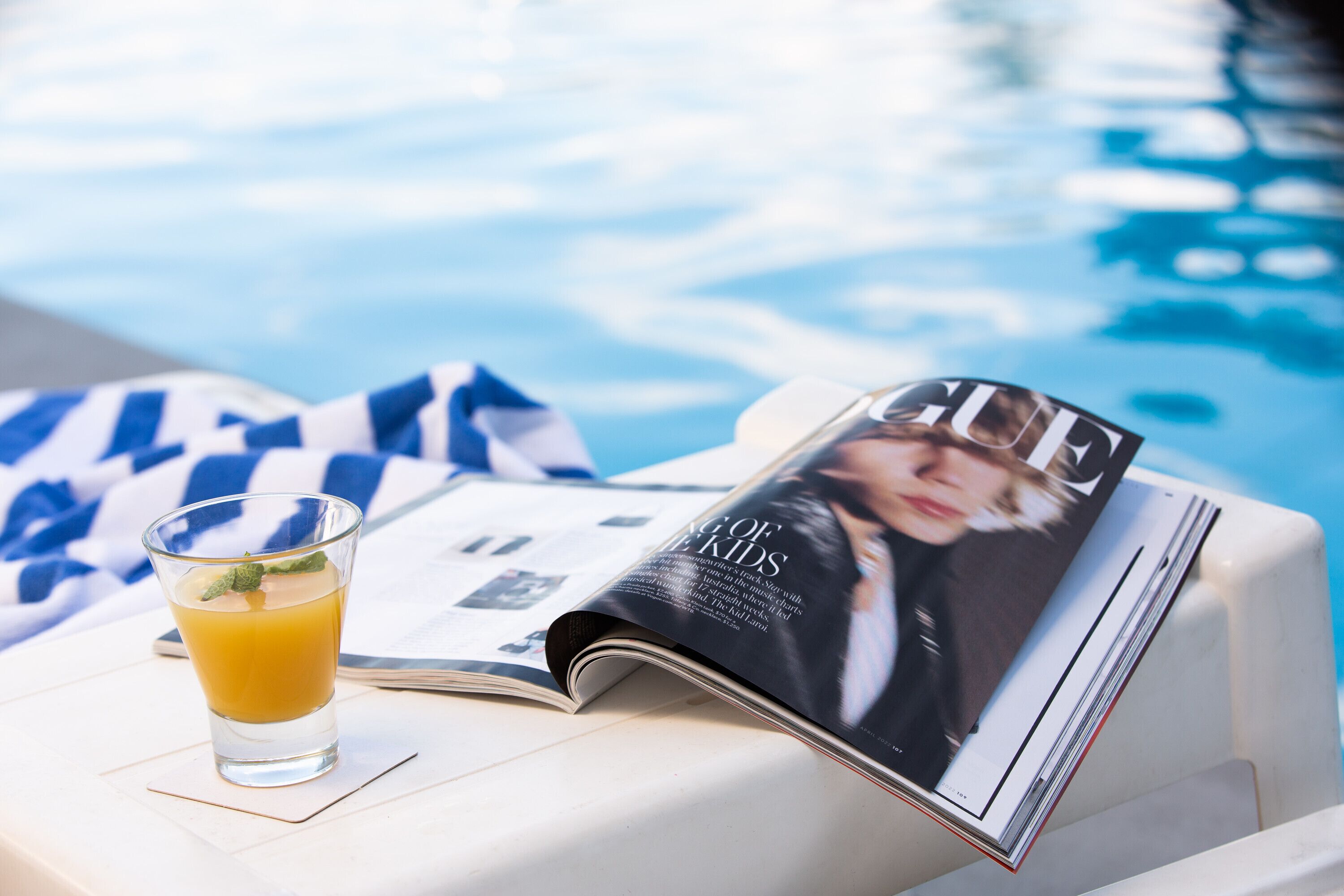 Revista em uma cadeira de lounge perto de uma piscina, com um copo de suco de laranja.