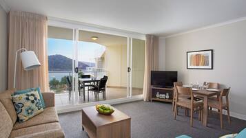 Uma sala de estar moderna com um sofá, uma mesa de centro com frutas, uma área de jantar com cadeiras e uma mesa, uma TV e uma grande porta de vidro deslizante que dá para uma varanda externa.