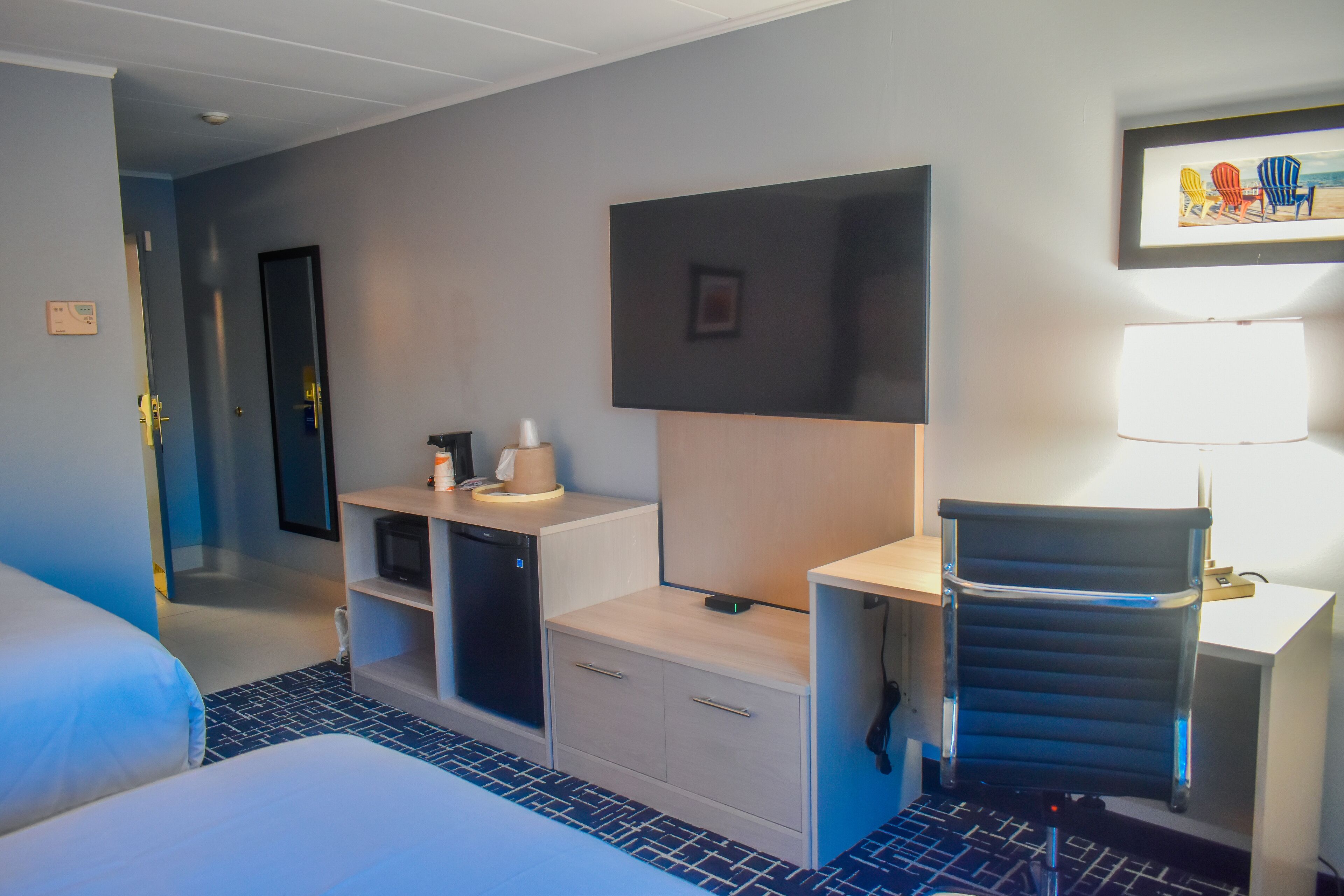 Quarto de hotel com cama, escrivaninha com cadeira, televisão, abajur e prateleira pequena com cafeteira.
