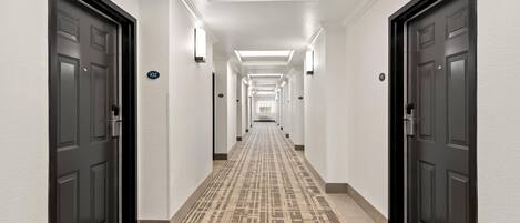 Corredor de hotel com portas escuras, paredes claras e carpete estampado.