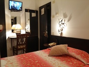 Quarto de hotel com uma cama, uma escrivaninha com abajur, uma cadeira, uma TV fixada na parede e uma porta com espelho.