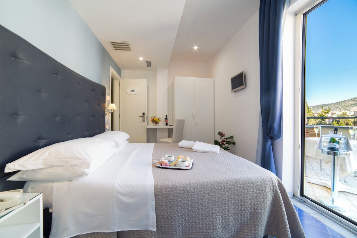 Quarto de hotel com uma cama grande, mesa de cabeceira, janela com vista para o exterior e banheiro visível através de uma porta aberta.
