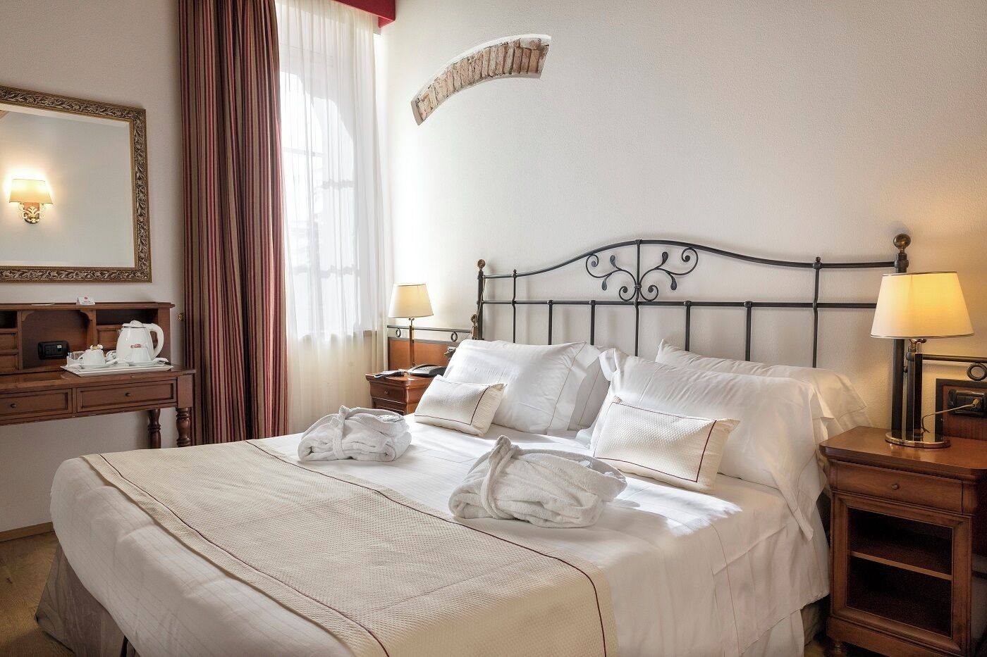 Quarto de hotel com uma cama grande, duas mesas de cabeceira, uma escrivaninha com telefone, um espelho e uma janela com cortinas.
