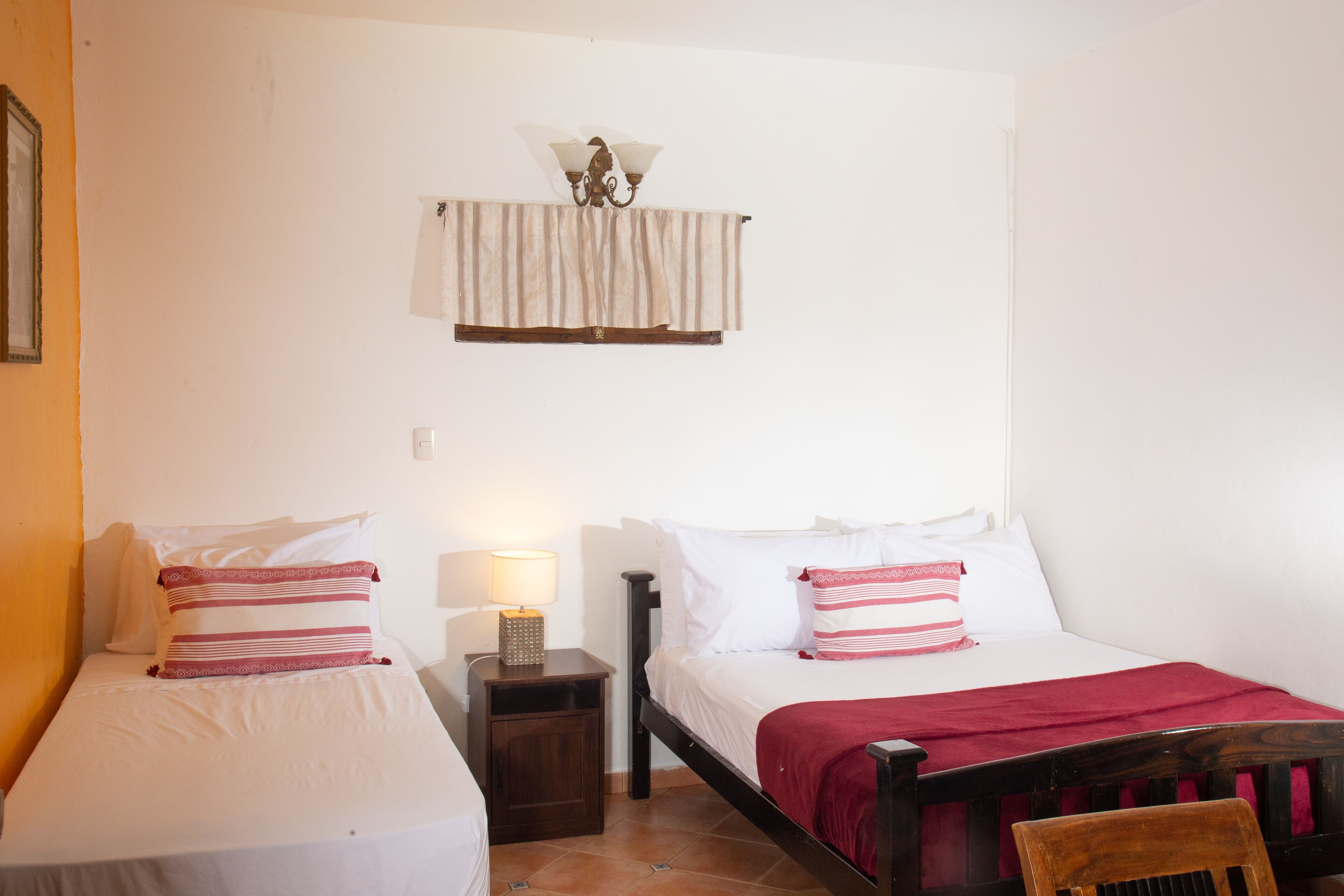 Quarto com duas camas, mesa de cabeceira com abajur e luminária na parede.