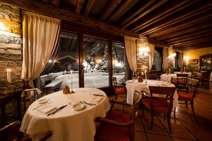 Restaurante com mesas redondas preparadas para refeições, paredes de pedra e vista para uma paisagem nevada através de grandes janelas.