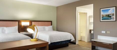 Quarto de hotel com duas camas, uma escrivaninha com candeeiro, mesa de cabeceira e banheiro visível por uma porta aberta.