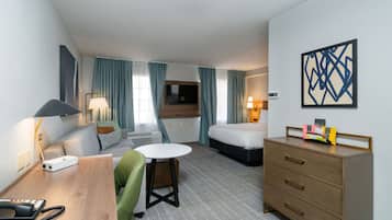 Quarto de hotel com cama, escrivaninha com luminária, cadeira, cômoda e televisão presa na parede.
