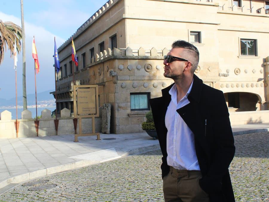 Um homem em frente a um edifício com fachada de pedra, bandeiras e chão de paralelepípedos.