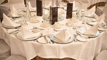 Uma sala de banquetes com mesas redondas preparadas para um evento formal, cada uma com toalhas brancas, talheres e copos.