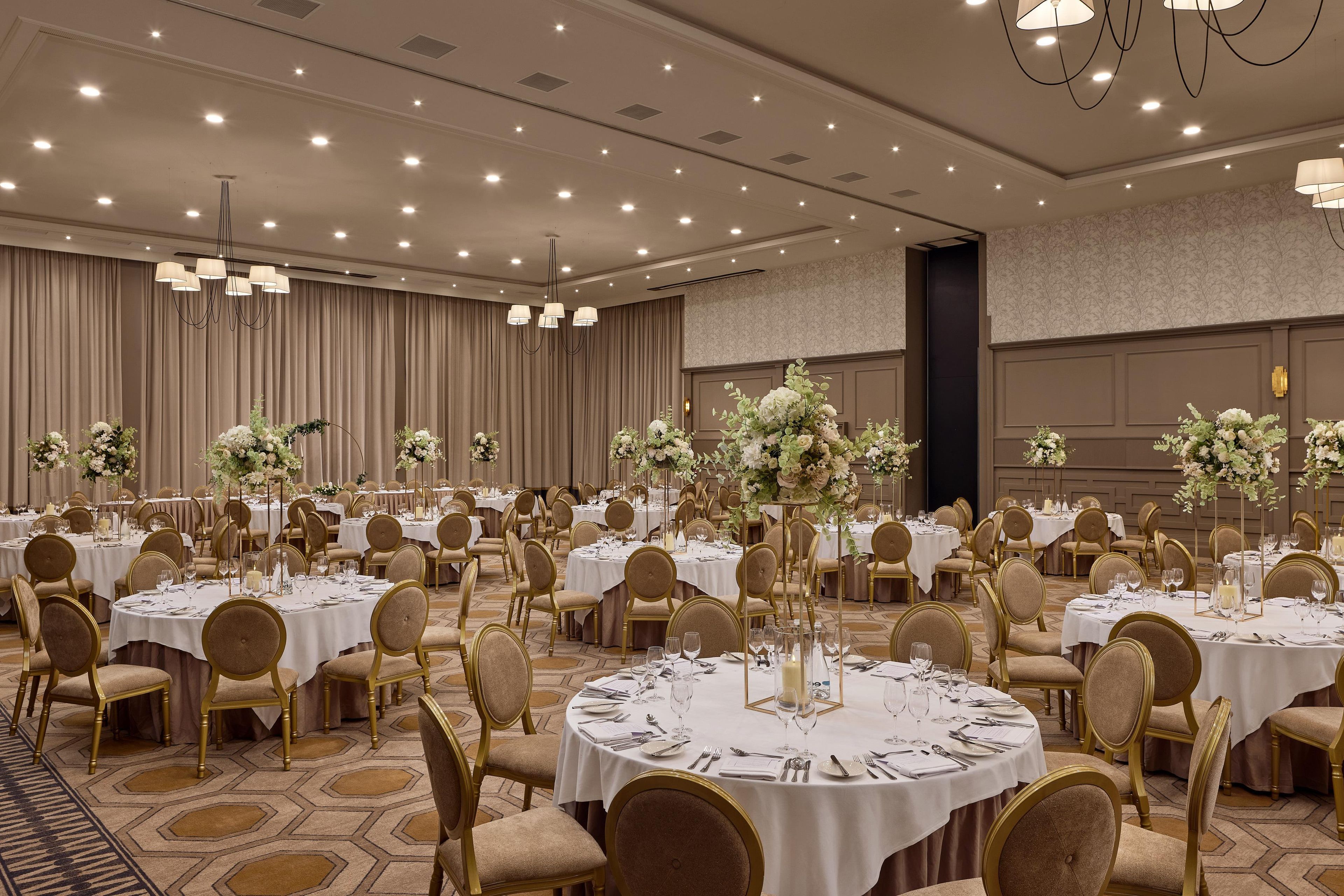 Uma sala de banquetes com mesas redondas, cadeiras douradas e arranjos florais.