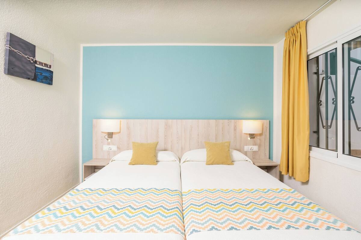 Uma cama de casal com colcha estampada em padrão ziguezague, duas luminárias de cabeceira, um painel de cabeceira de madeira e uma janela com cortinas amarelas.
