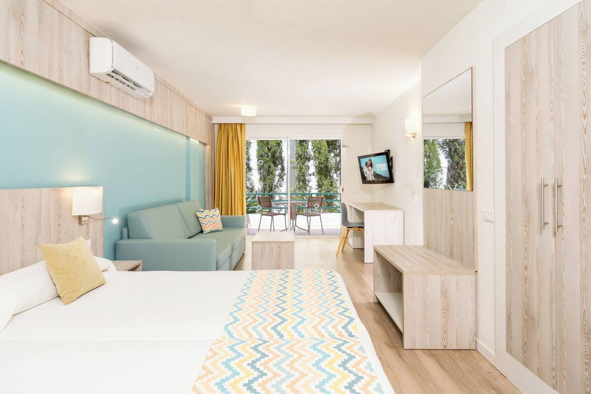 Quarto de hotel moderno com cama, sofá, escrivaninha e varanda com vista para árvores.