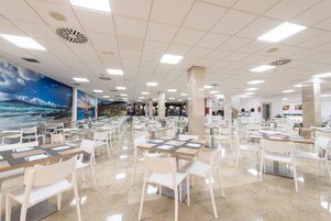 Uma cafeteria ampla com várias mesas e cadeiras, um mural na parede e um chão limpo revestido de azulejos.
