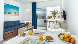 Quarto de hotel moderno com sofá, mesa de café da manhã e varanda com vista para palmeiras.